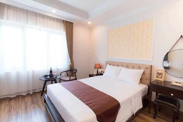 Quarto de hotel com uma cama grande, uma escrivaninha, uma cadeira, uma mesa de cabeceira com abajur e uma janela com cortinas.