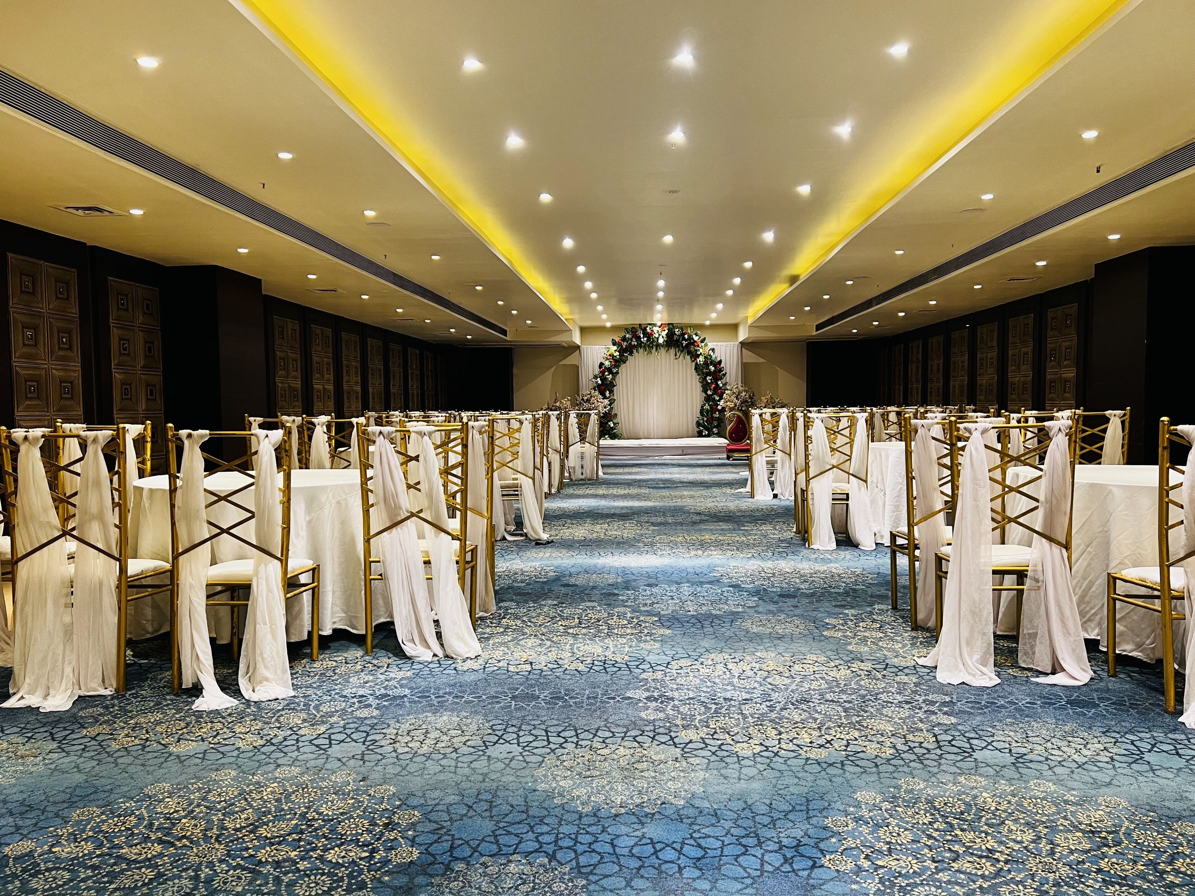 Uma sala de banquetes com fileiras de mesas e cadeiras, um tapete estampado em azul e um palco com um arco floral.