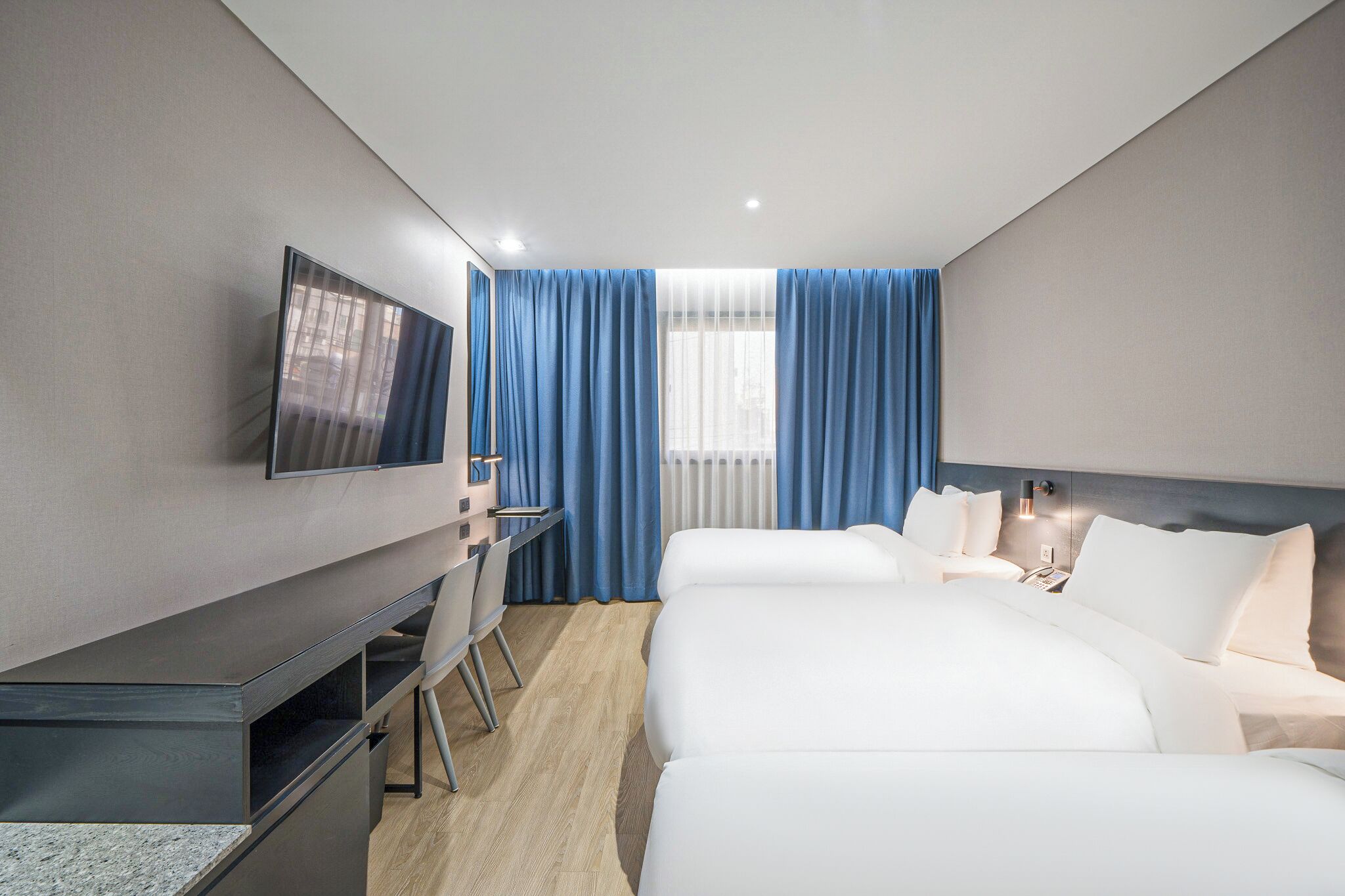 Quarto de hotel com uma cama, uma escrivaninha com cadeira, televisão e uma janela com cortinas.