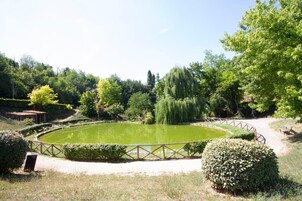 Um lago circular com cerca de madeira, rodeado por arbustos e árvores bem cuidados.