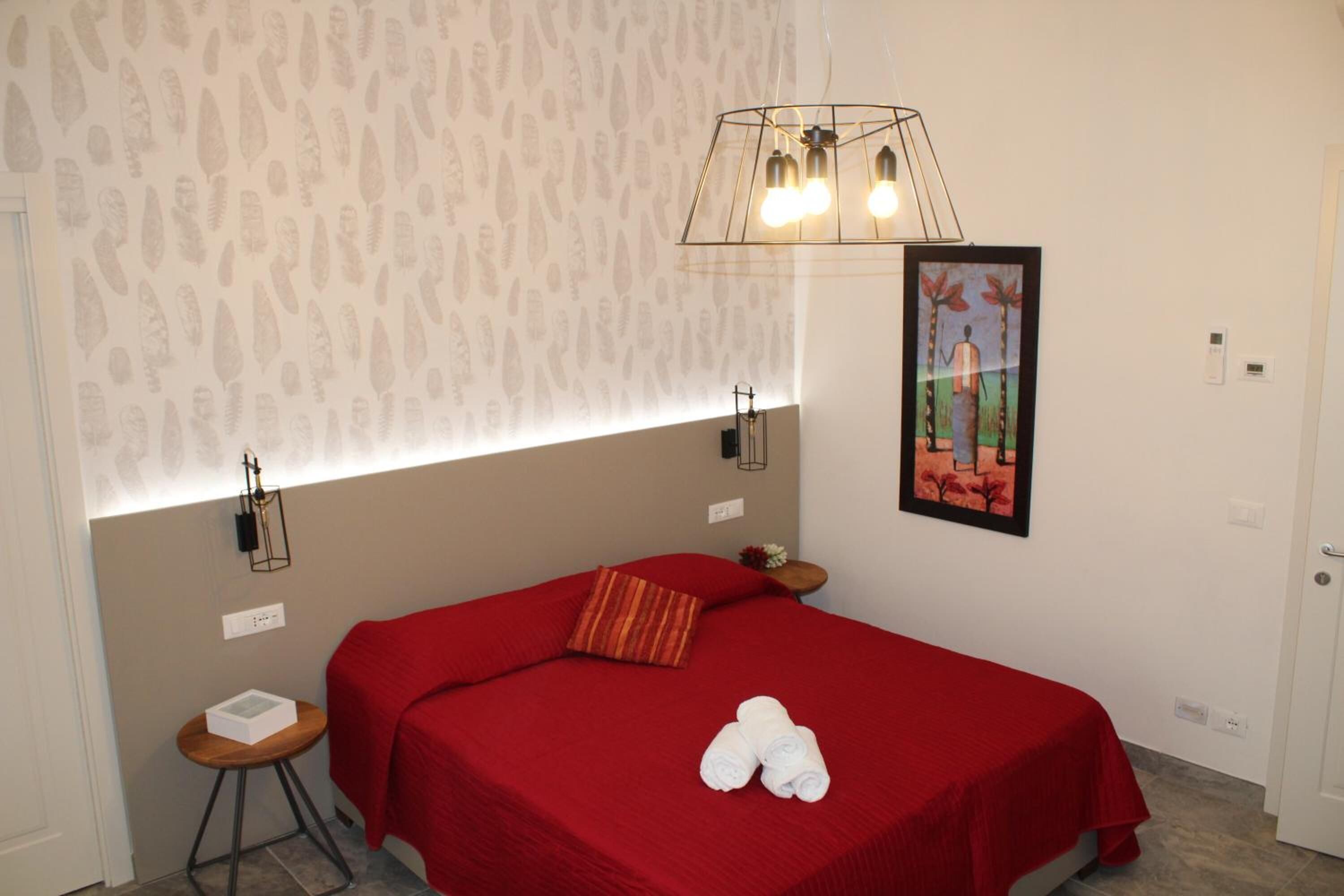 Quarto de hotel com cama vermelha, um banquinho de madeira, luminária fixada na parede e uma moldura com um quadro.