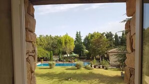 Visão através de uma janela emoldurada em pedra de um quintal com piscina, gramado verde e diversas árvores.