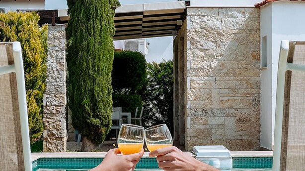 Vista da piscina com duas pessoas brindando com bebidas, uma parede de pedra e um telhado telhado.
