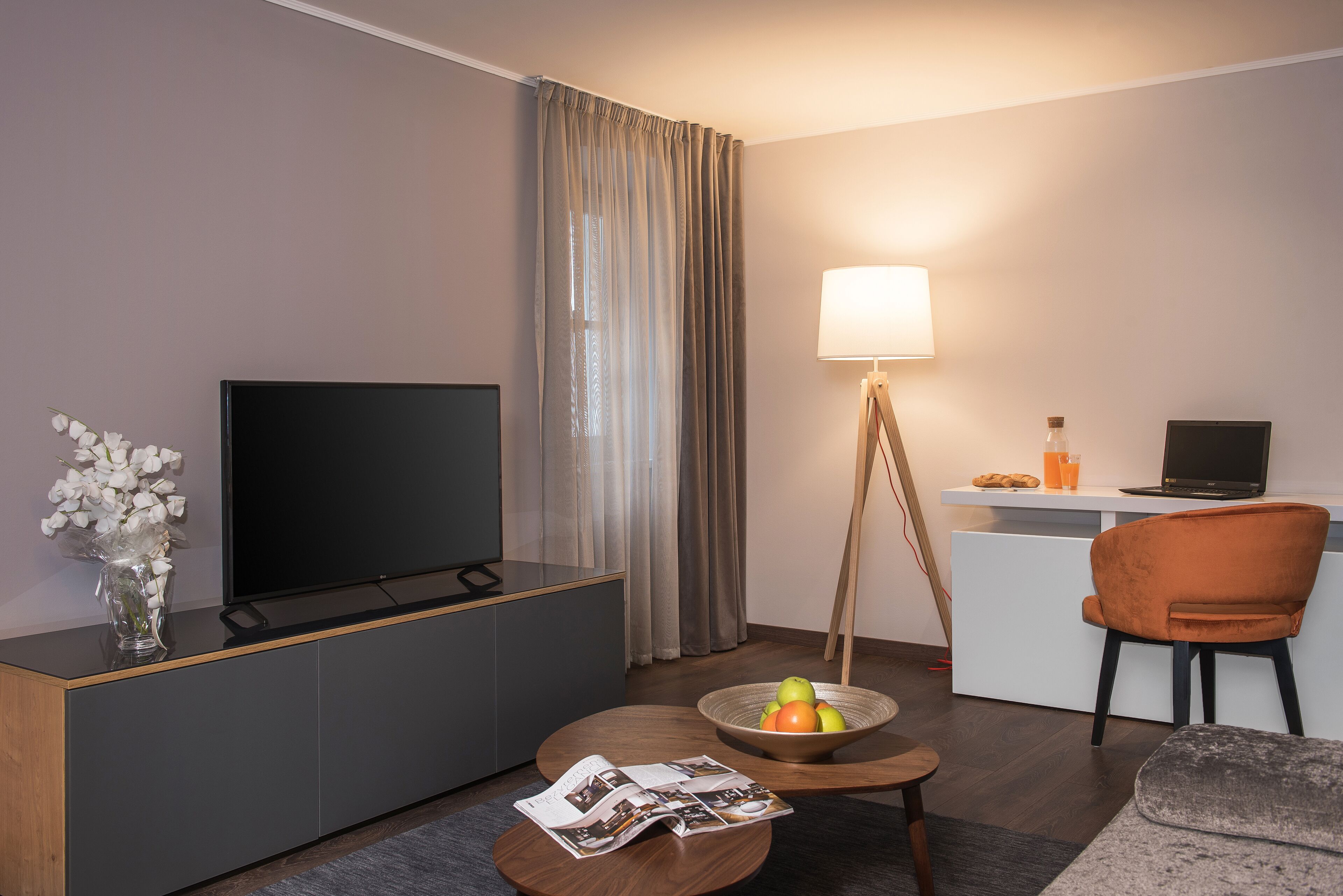 Quarto de hotel moderno com TV de tela plana, uma escrivaninha com um laptop, uma cadeira, uma mesinha de centro com uma revista e uma cesta de frutas.
