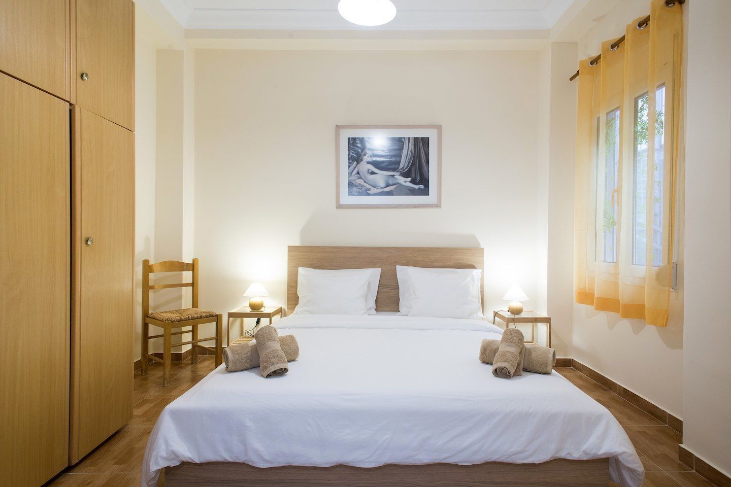 Quarto de hotel com uma cama grande, duas mesas de cabeceira com abajures, uma cadeira de madeira, um guarda-roupa e um quadro na parede.