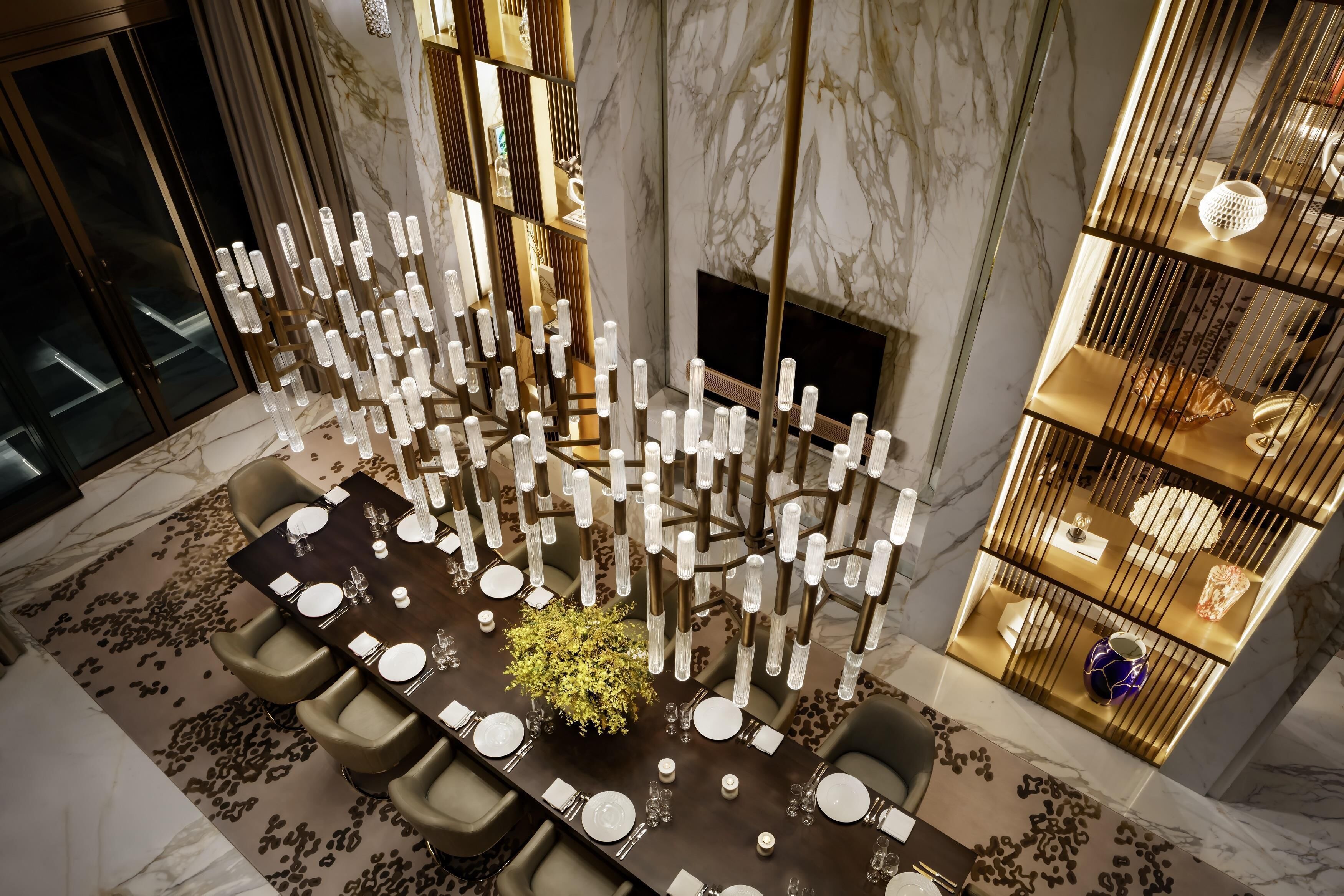 Uma sala de jantar com uma mesa comprida, várias cadeiras, luminárias altas em formato de vela e um arranjo de plantas decorativo.