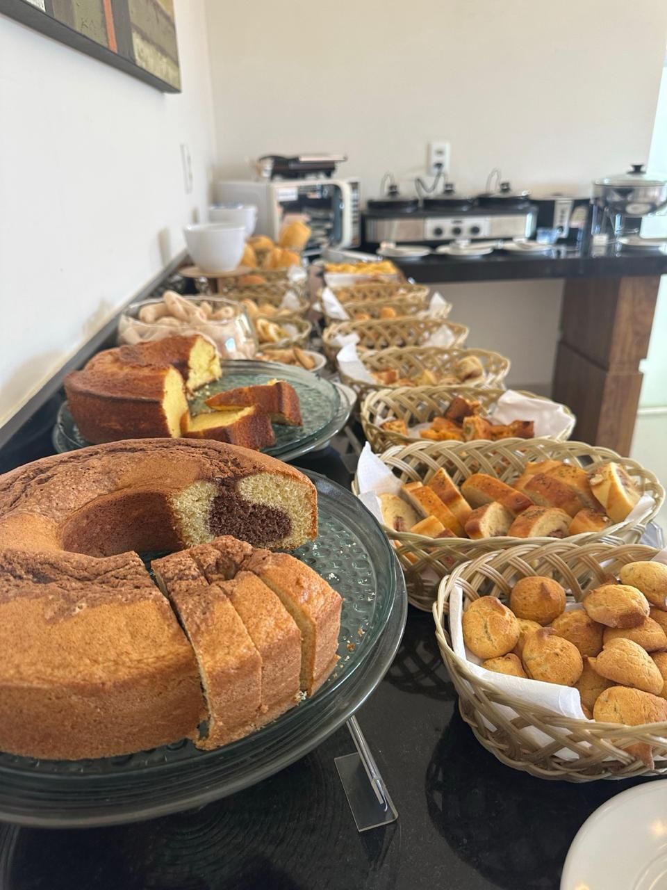 Uma mesa de buffet com diversos produtos de panificação, incluindo um bolo bundt grande, muffins e doces.
