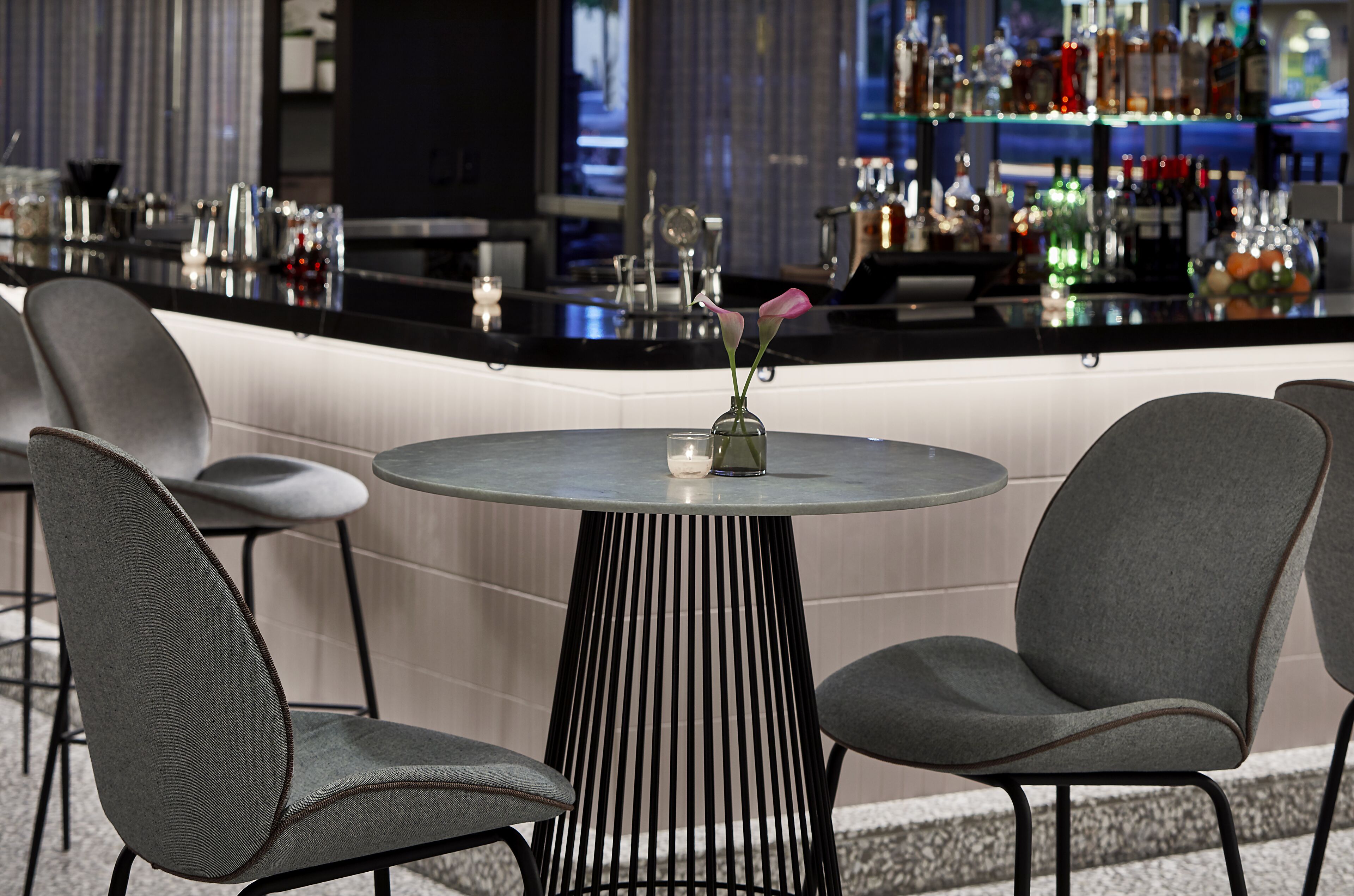 Um espaço de bar com uma mesa redonda, duas cadeiras cinzentas e um vaso com flores.