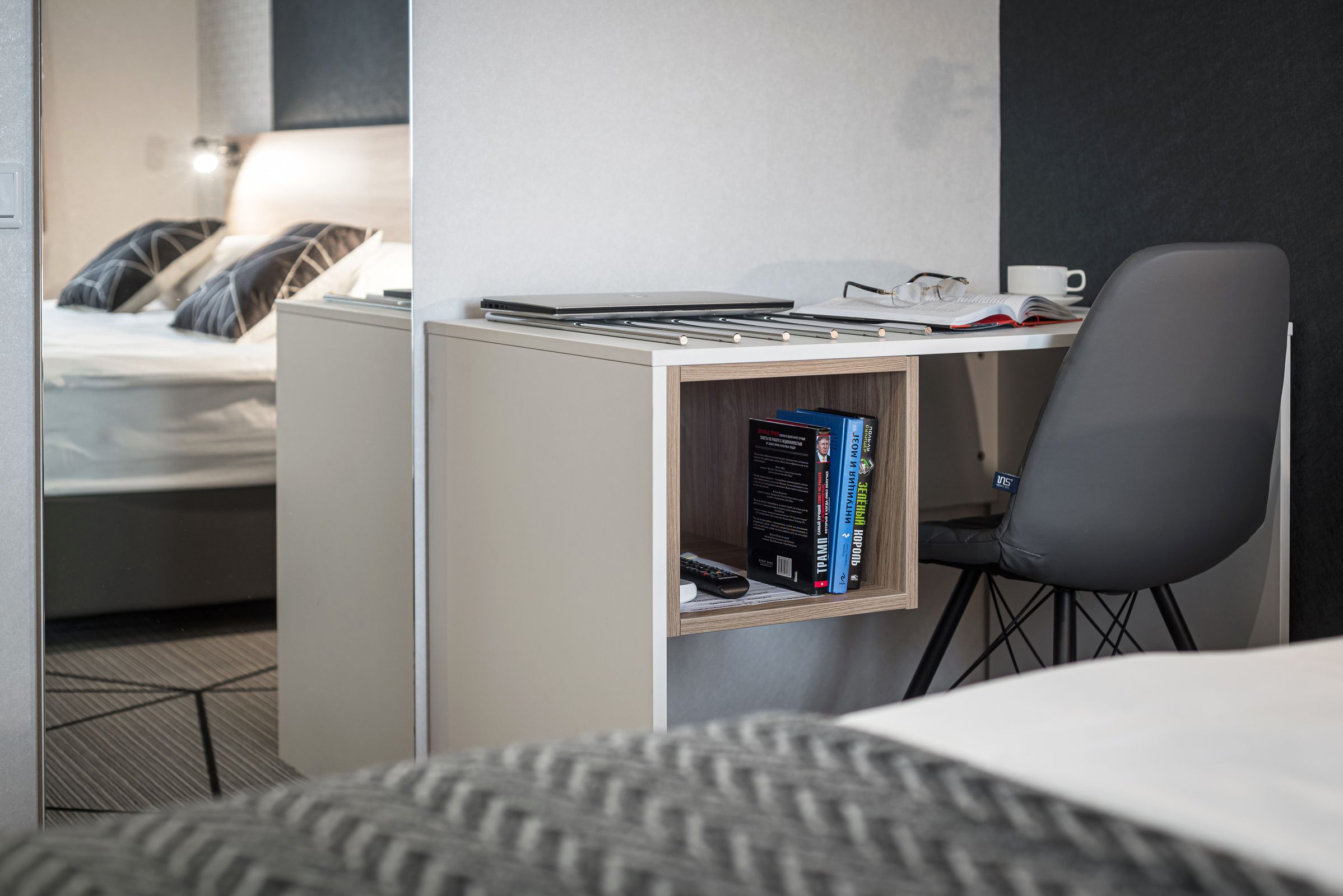 Quarto de hotel compacto com uma pequena escrivaninha, uma cadeira, uma cama e uma estante de livros.
