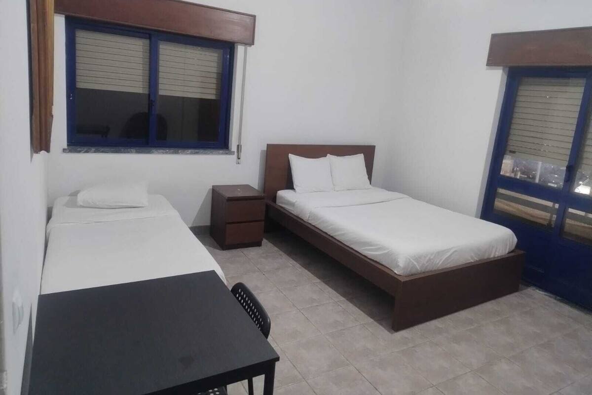 Quarto pequeno e limpo, com duas camas individuais, uma mesinha e janela com persianas.