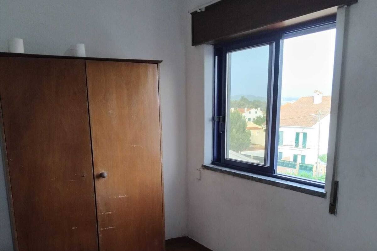 Quarto com armário de madeira, cama com colchão branco e janela com vista para prédios.