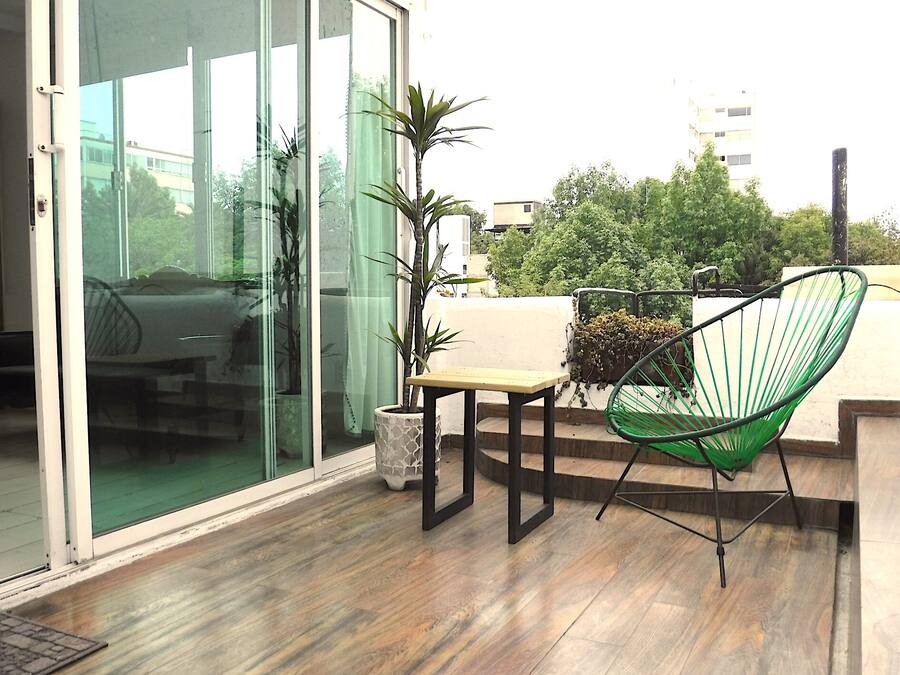 Uma varanda com piso de madeira, uma cadeira verde, uma mesa pequena e um vaso com planta.