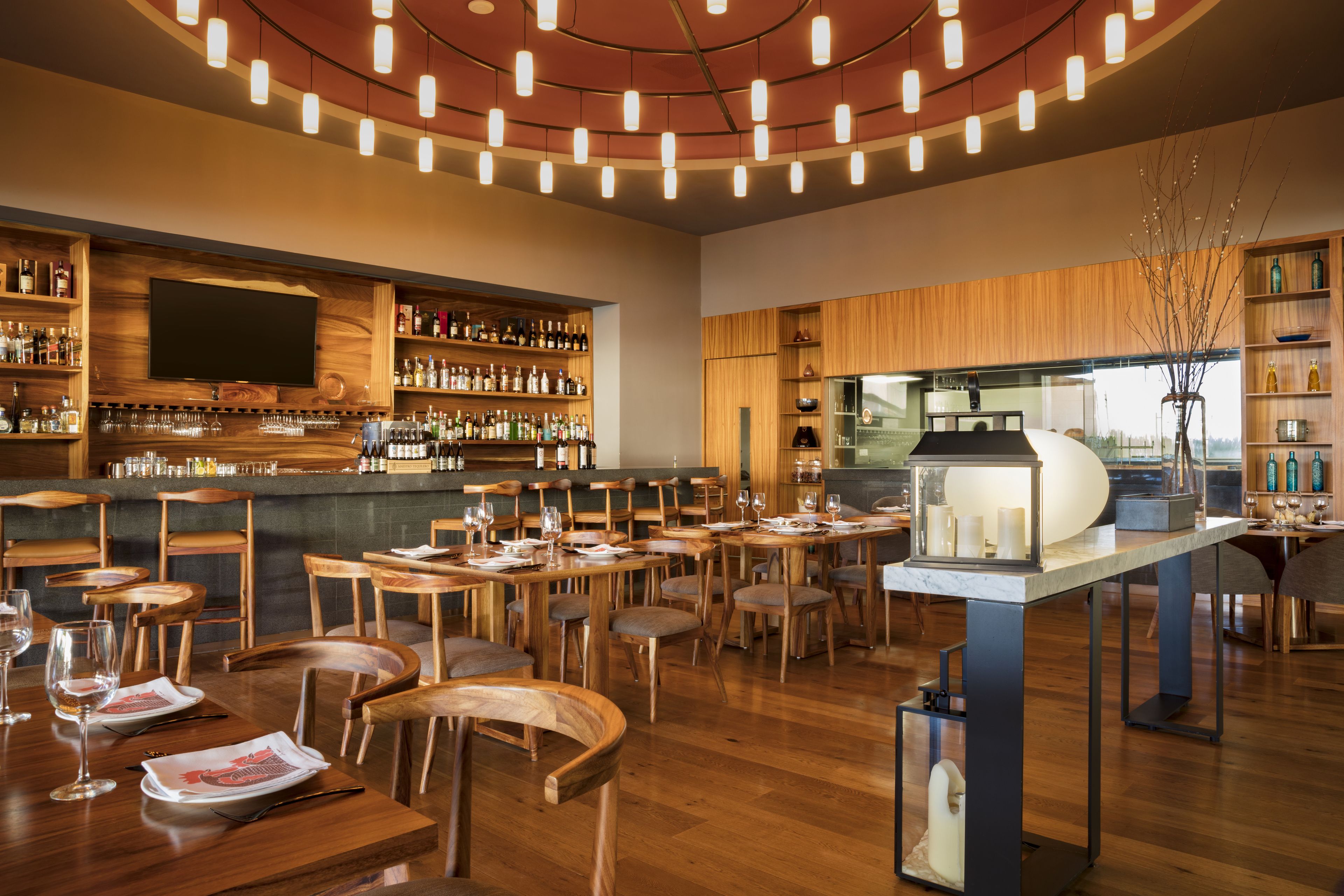 Um restaurante com mesas e cadeiras de madeira, área de bar com prateleiras cheias de garrafas e uma grande tela de televisão.
