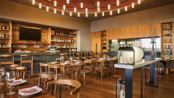 Um restaurante com mesas e cadeiras de madeira, área de bar com prateleiras cheias de garrafas e uma grande tela de televisão.