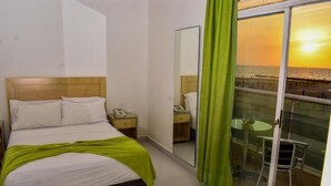 Quarto de hotel com cama, mesa de cabeceira, espelho e varanda com vista para o pôr do sol.