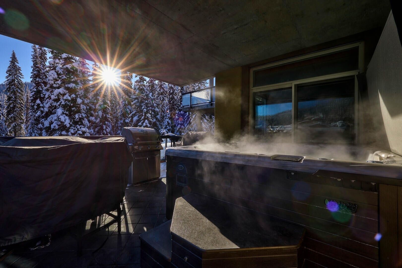 Uma área de cozinha externa com churrasqueira e banheira de hidromassagem, cercada por árvores cobertas de neve e um céu claro.