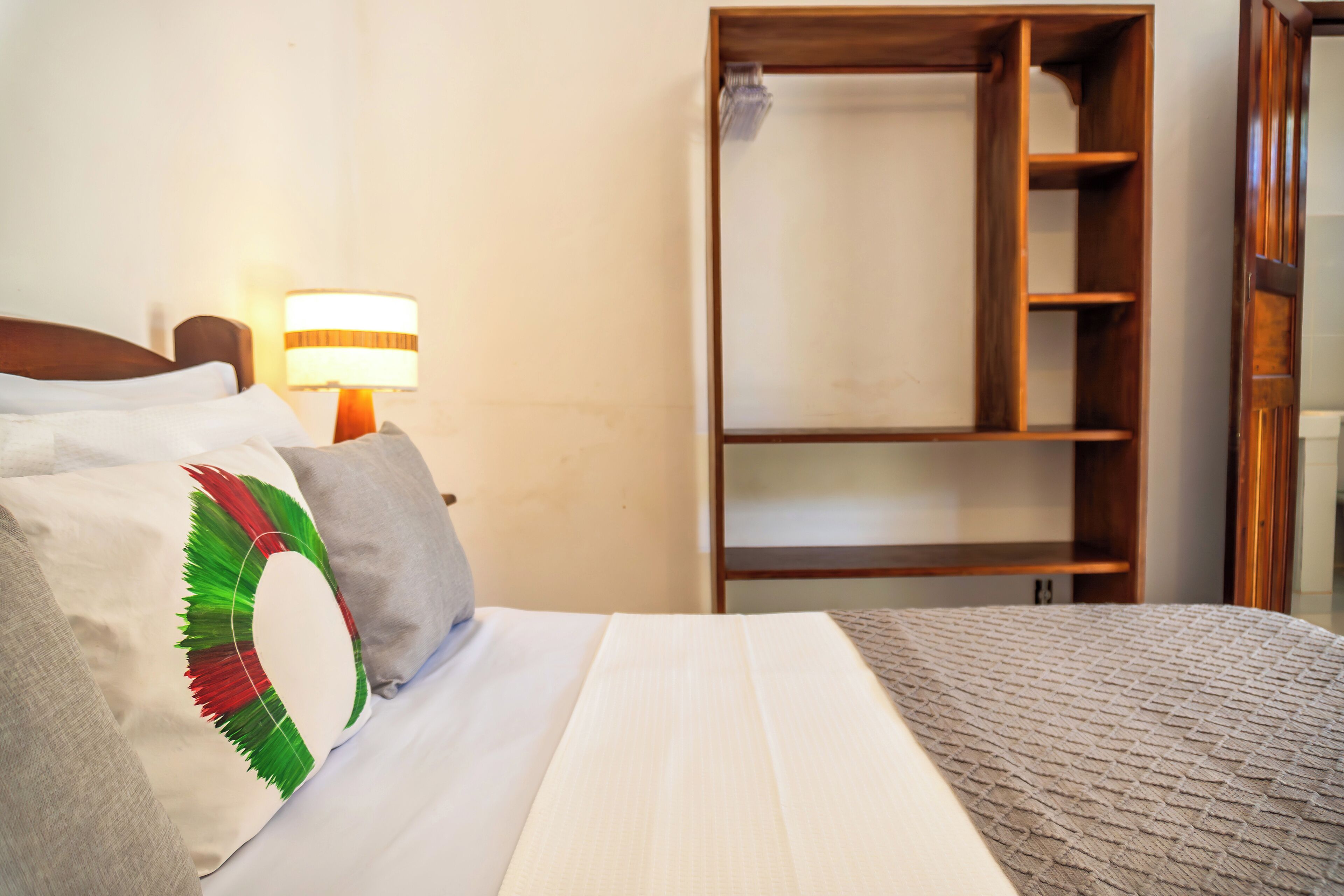 Uma cama bem arrumada com um cobertor branco e cinza, um travesseiro com desenho verde e vermelho, e um guarda-roupa de madeira com portas abertas.