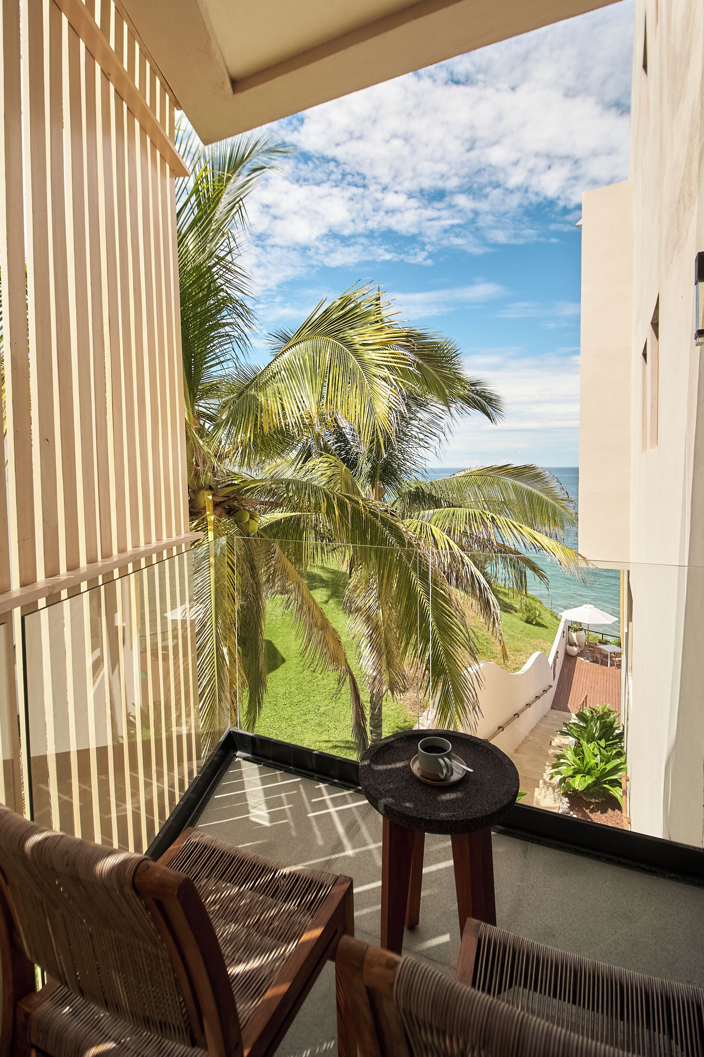 Uma varanda com vista para o oceano, uma palmeira e uma pequena mesa com uma xícara.
