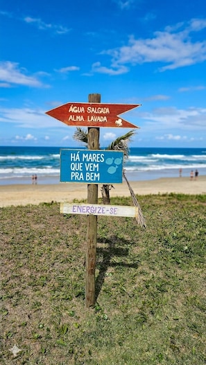 Placa de praia com várias setas indicativas e texto, cercada por uma área gramada e o oceano ao fundo.