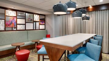 Um hall de hotel moderno com uma mesa comprida, cadeiras estofadas em azul, um banquinho vermelho e um sofá verde.