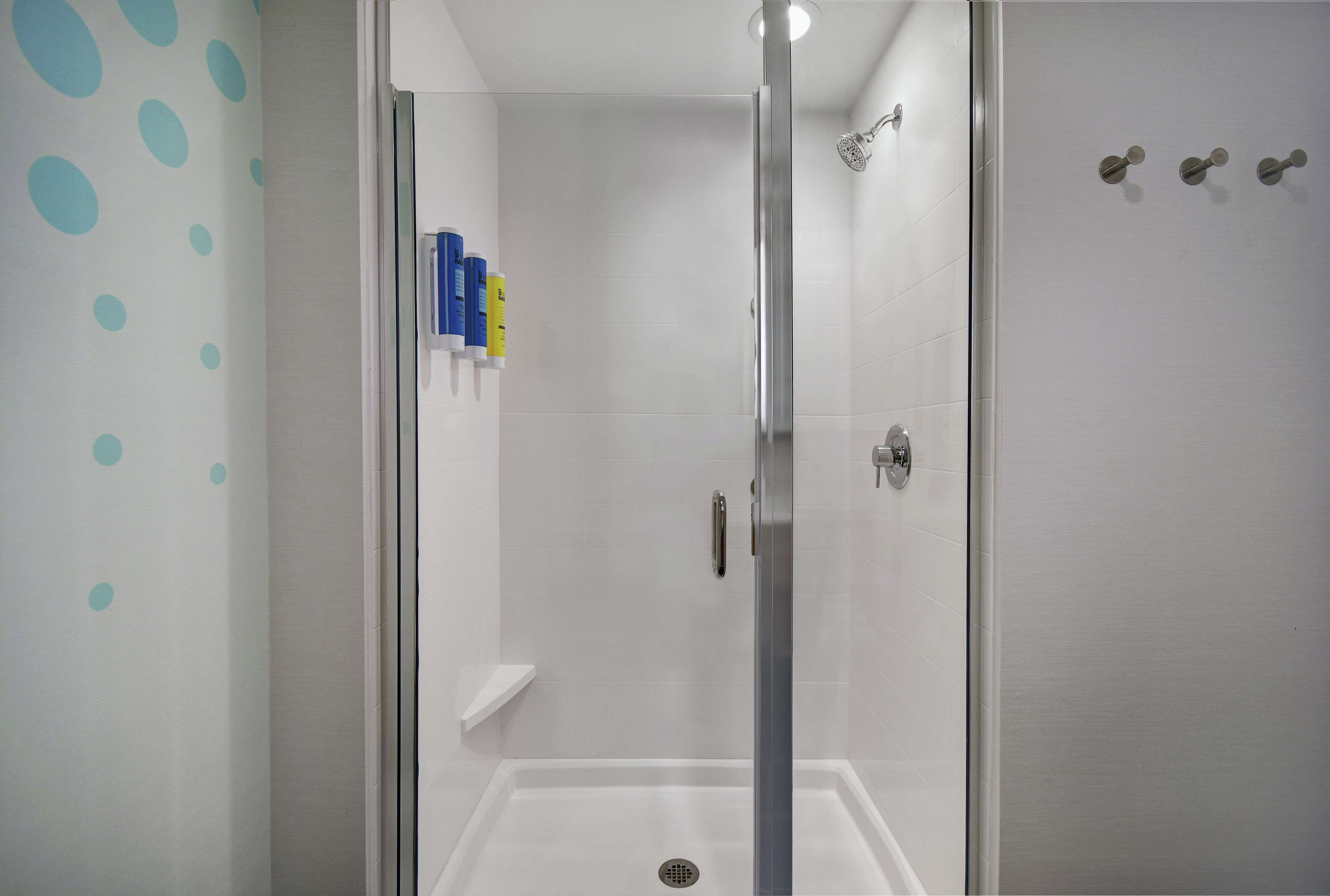 Divisória de chuveiro envidraçada com parede de azulejos brancos, chuveiro e prateleira com produtos de higiene pessoal.