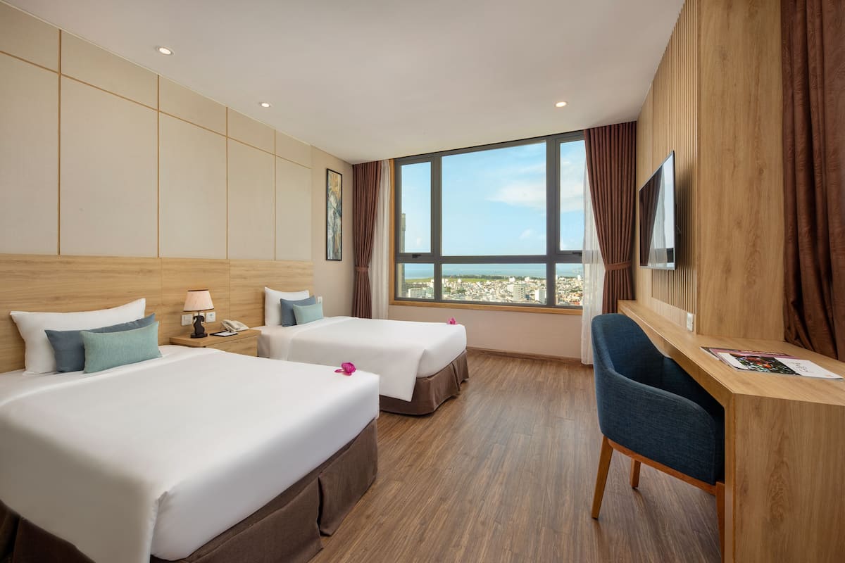 Quarto de hotel com duas camas, uma escrivaninha e uma janela grande com vista para a cidade.