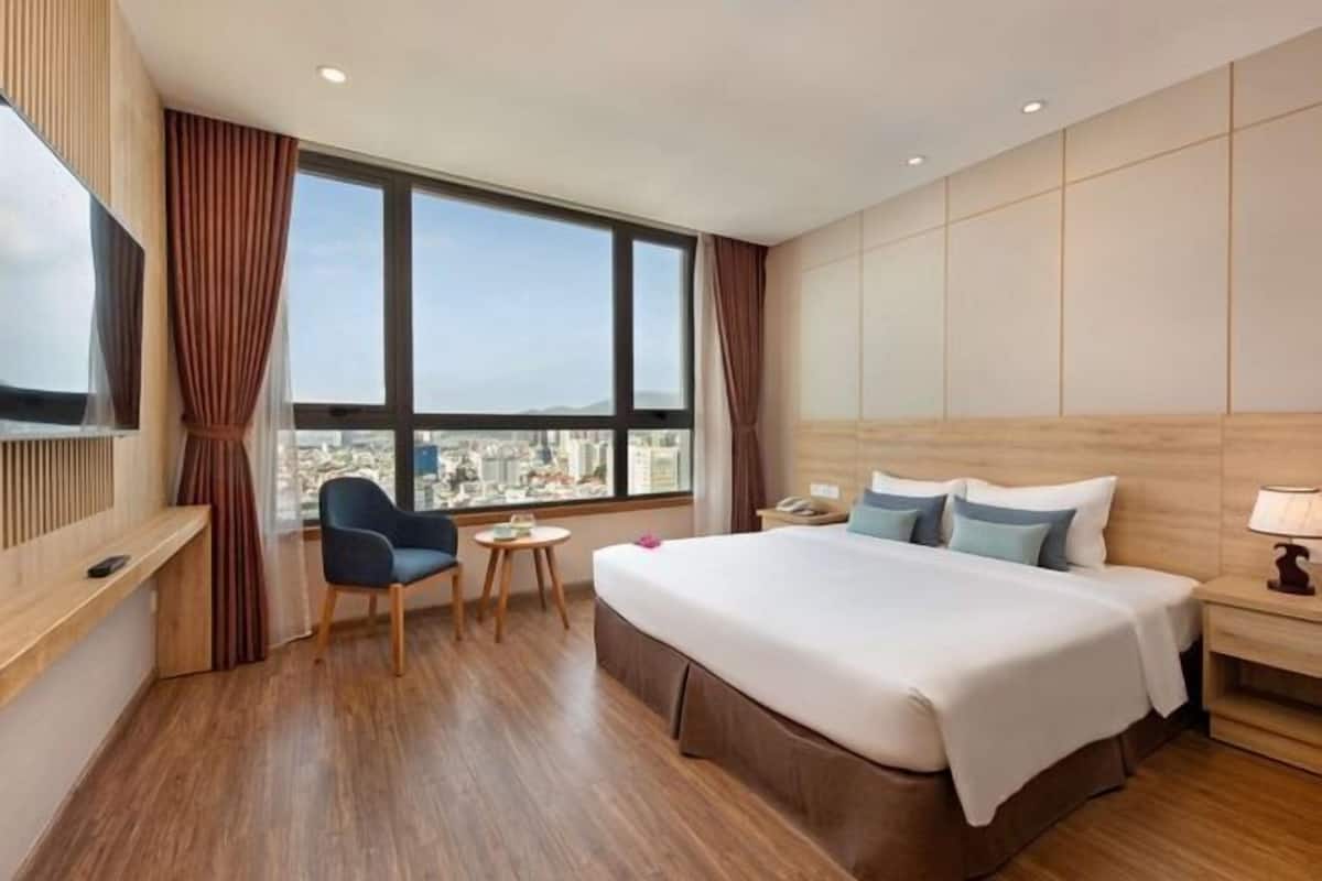 Quarto de hotel com uma cama grande, uma escrivaninha, uma cadeira, uma mesa pequena e uma janela ampla com vista para a cidade.