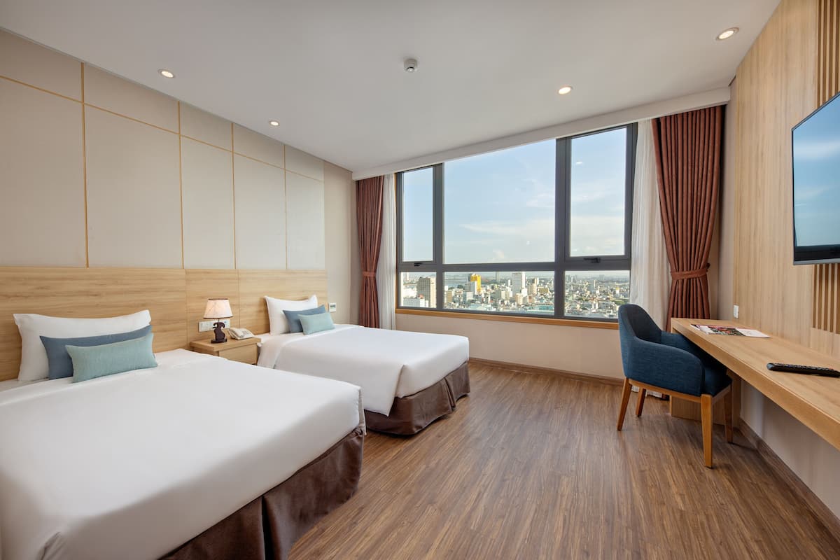 Quarto de hotel com duas camas, uma escrivaninha, uma cadeira, uma televisão e uma janela grande com vista para a cidade.