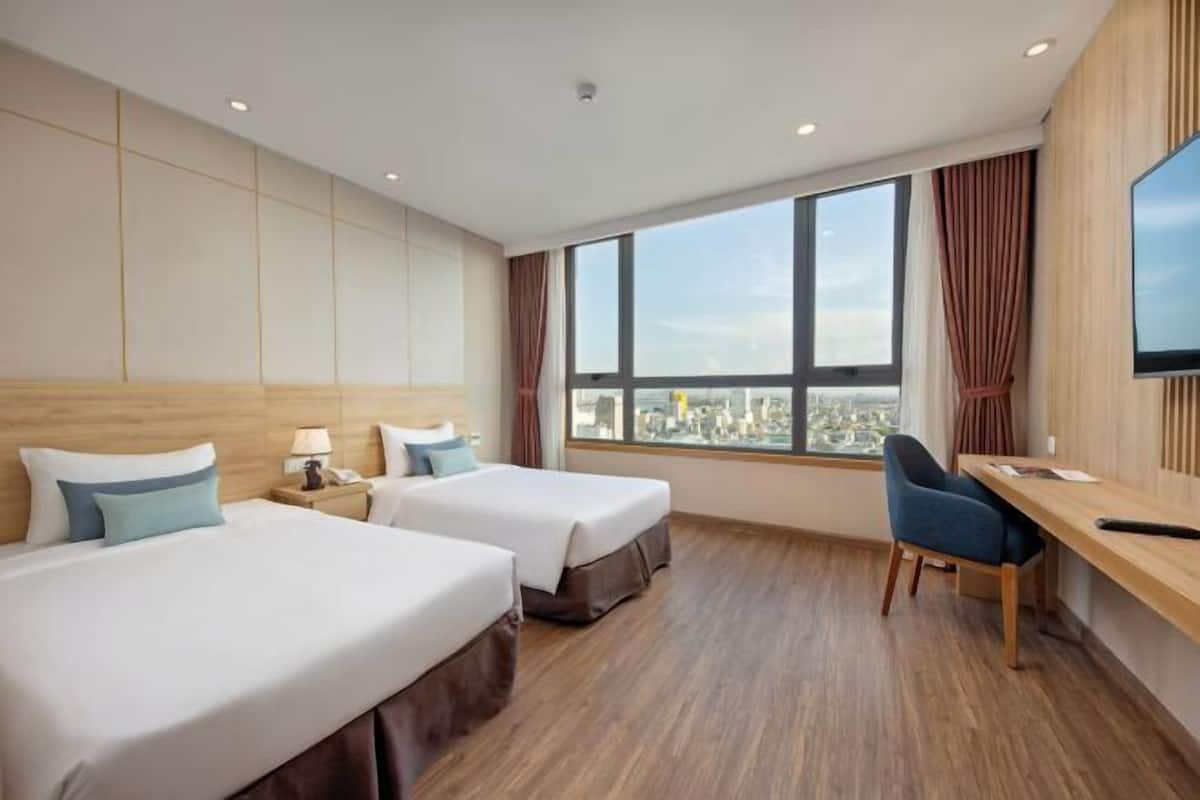 Quarto de hotel com duas camas, uma escrivaninha, uma cadeira, uma televisão e uma janela grande com vista para a cidade.