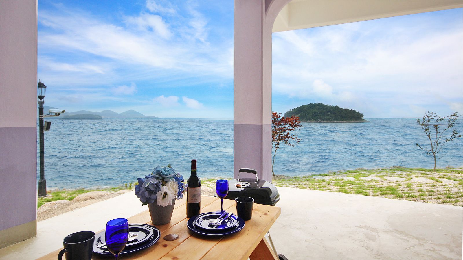 Mesa posta para dois, com garrafa de vinho, copos e um vaso de flores, com vista para um corpo d'água e uma ilha ao longe.
