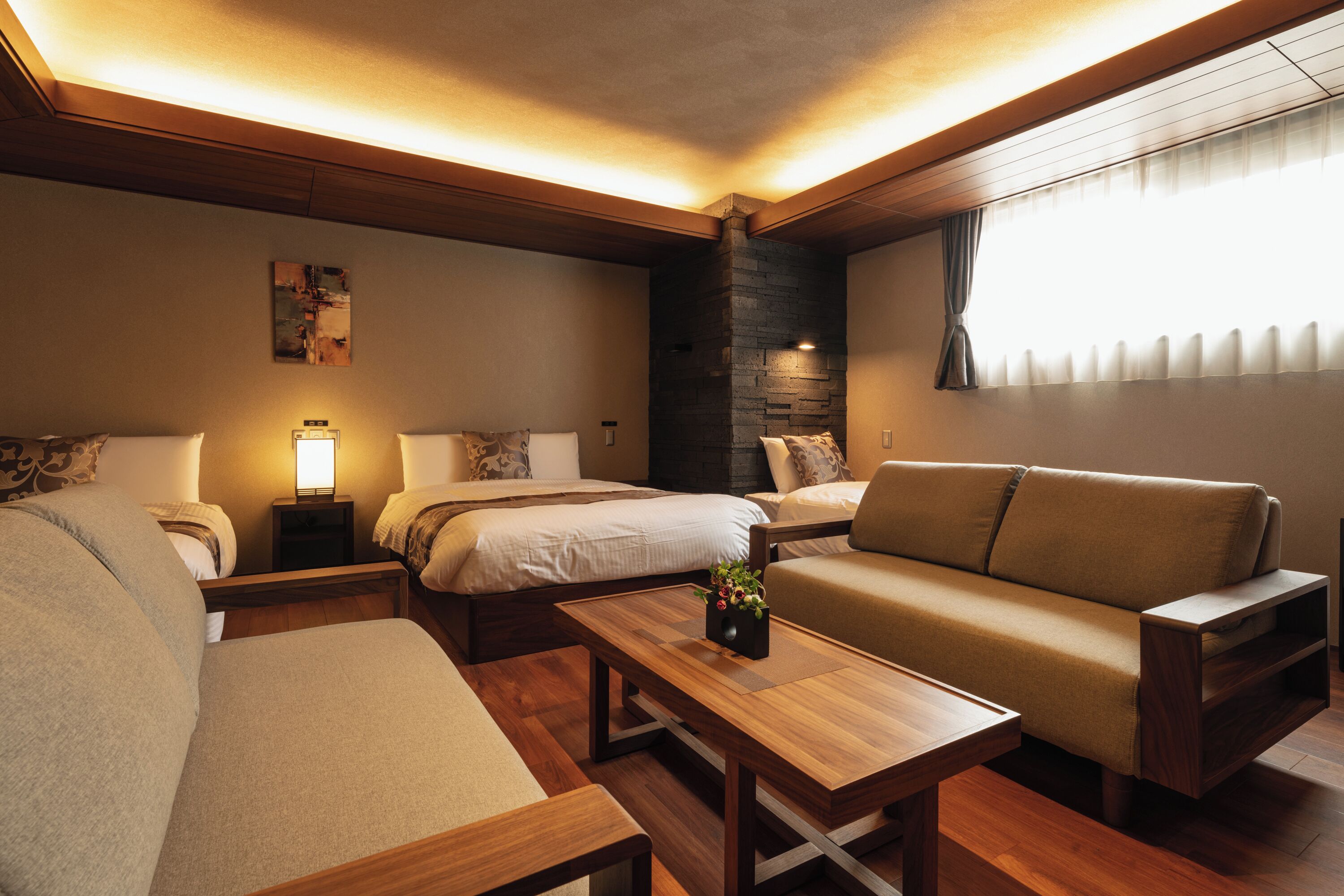 Quarto de hotel com uma cama, dois sofás, uma mesa de centro de madeira, uma mesa de cabeceira com abajur e uma parede de destaque em pedra.