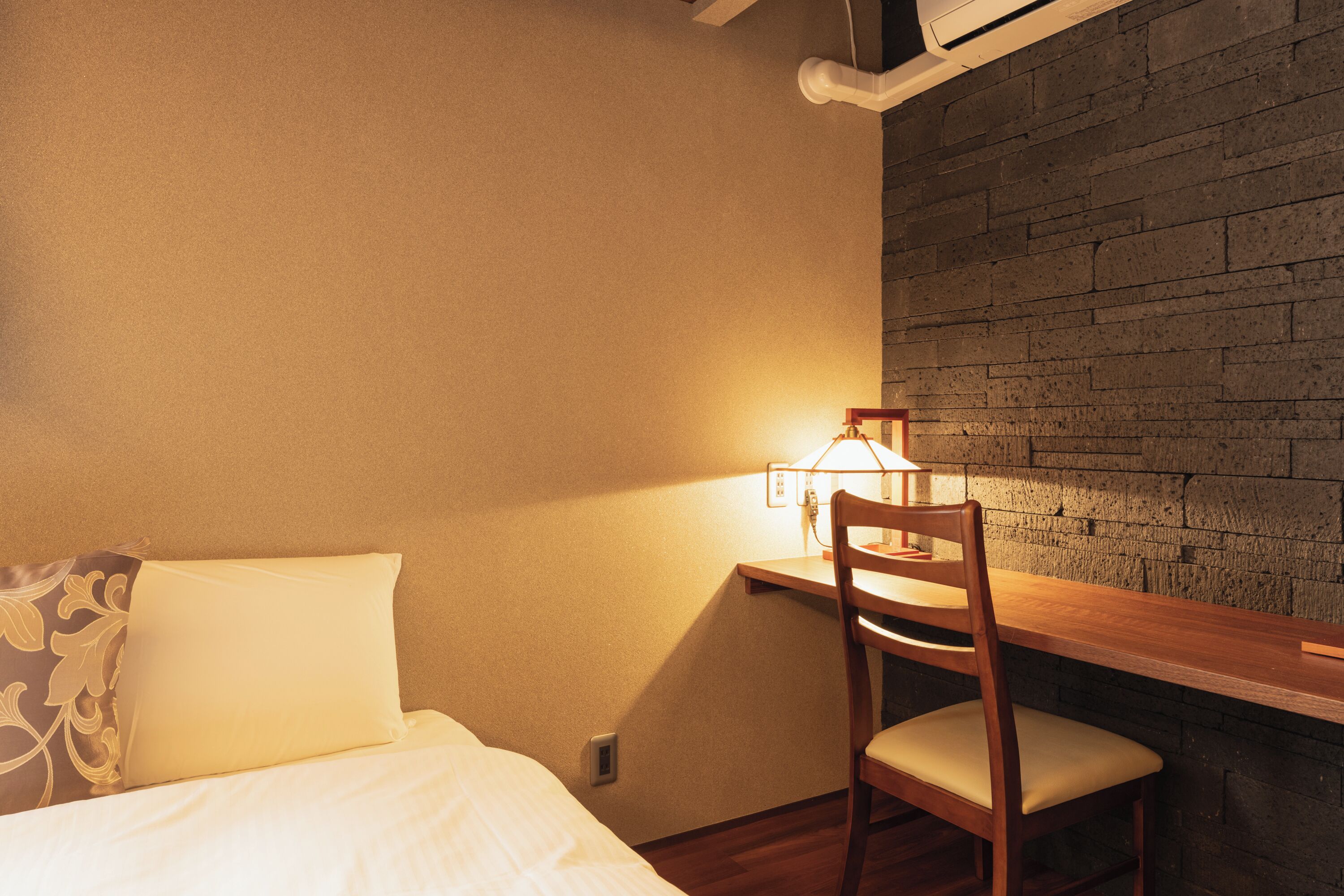 Quarto de hotel com cama, secretária de madeira, cadeira e luminária na parede.