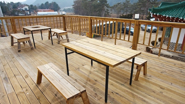 Um deck de madeira com mesas para piquenique e bancos, cercado por um corrimão de madeira, com vista para uma área residencial com casas e um edifício tradicional com telhado verde.