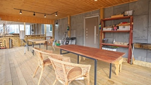 Interior em madeira com mesa e cadeiras de jantar, área de cozinha com prateleiras e vista pela janela.