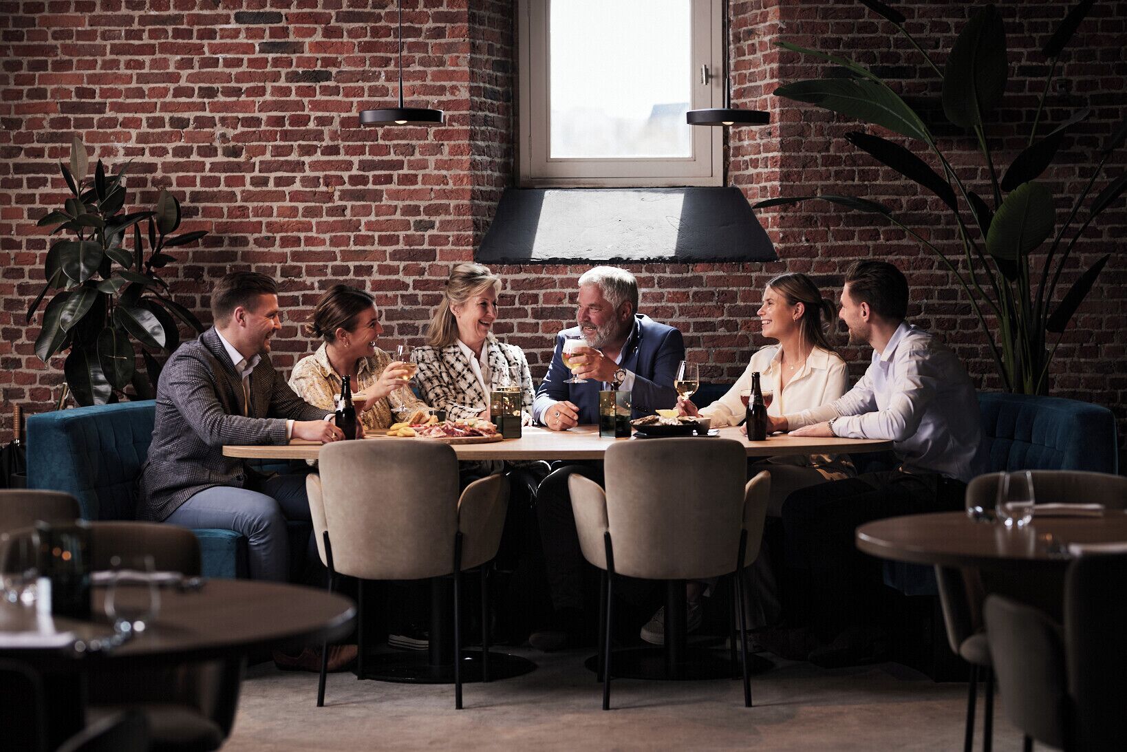 Um grupo de pessoas jantando à mesa em um salão com paredes de tijolo e uma janela, cercado por cadeiras e mesas.