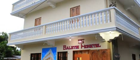 Um edifício de três andares com varandas, uma placa com a inscrição "Balefish Honavel" e uma pequena área de entrada com uma porta e um banco.