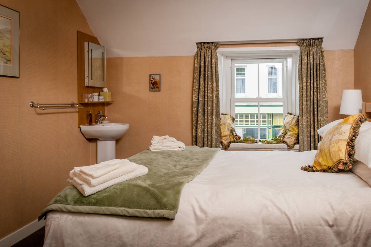 Quarto de hotel com cama, mesa de cabeceira, luminária, janela com cortinas, pia e espelho.