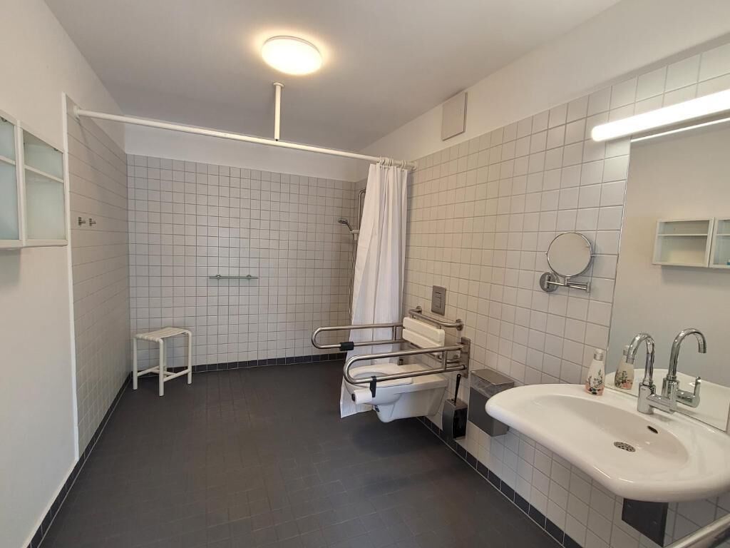 Casa de banho com lavatório branco, espelho, chuveiro com cortina branca e barra de apoio.