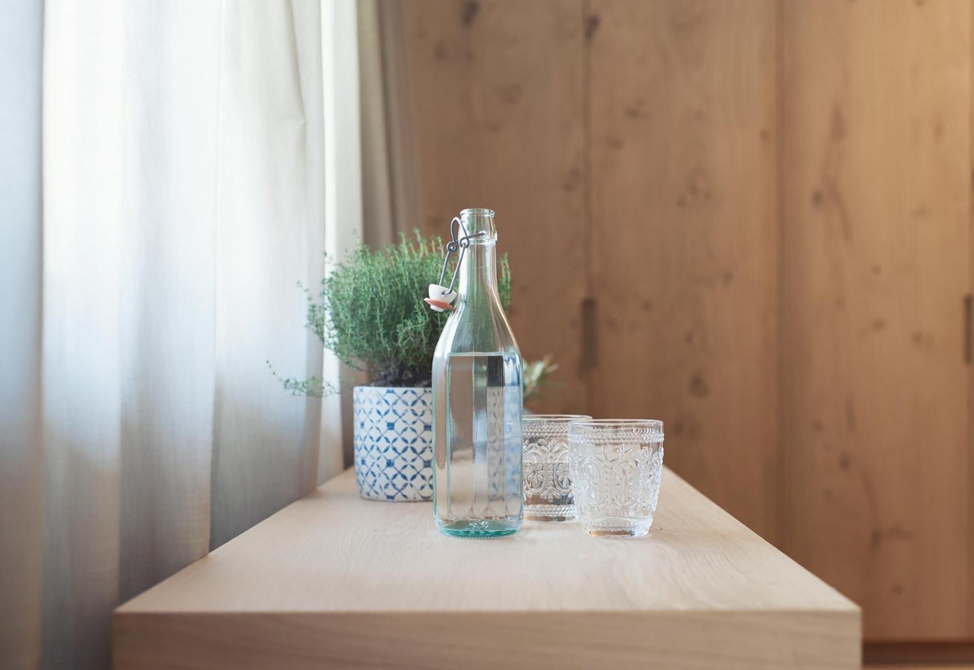 Uma garrafa de vidro transparente e duas taças de vidro sobre uma mesa de madeira, com uma planta em vaso ao fundo.