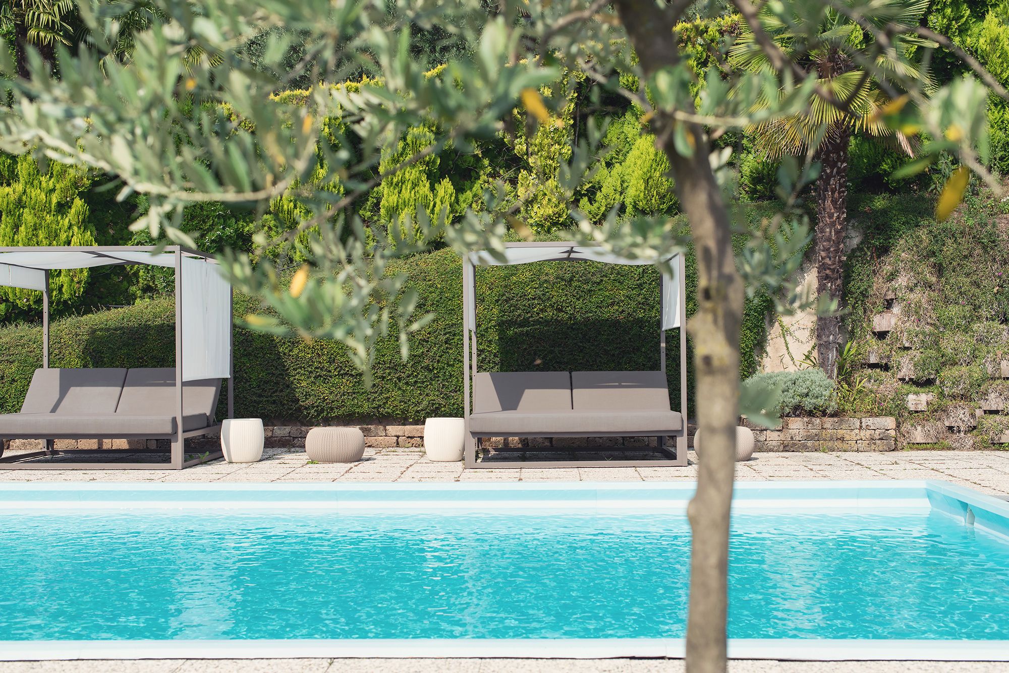 Uma área junto à piscina com duas espreguiçadeiras sob uma pérgula, rodeada por vegetação exuberante e um muro de pedra.