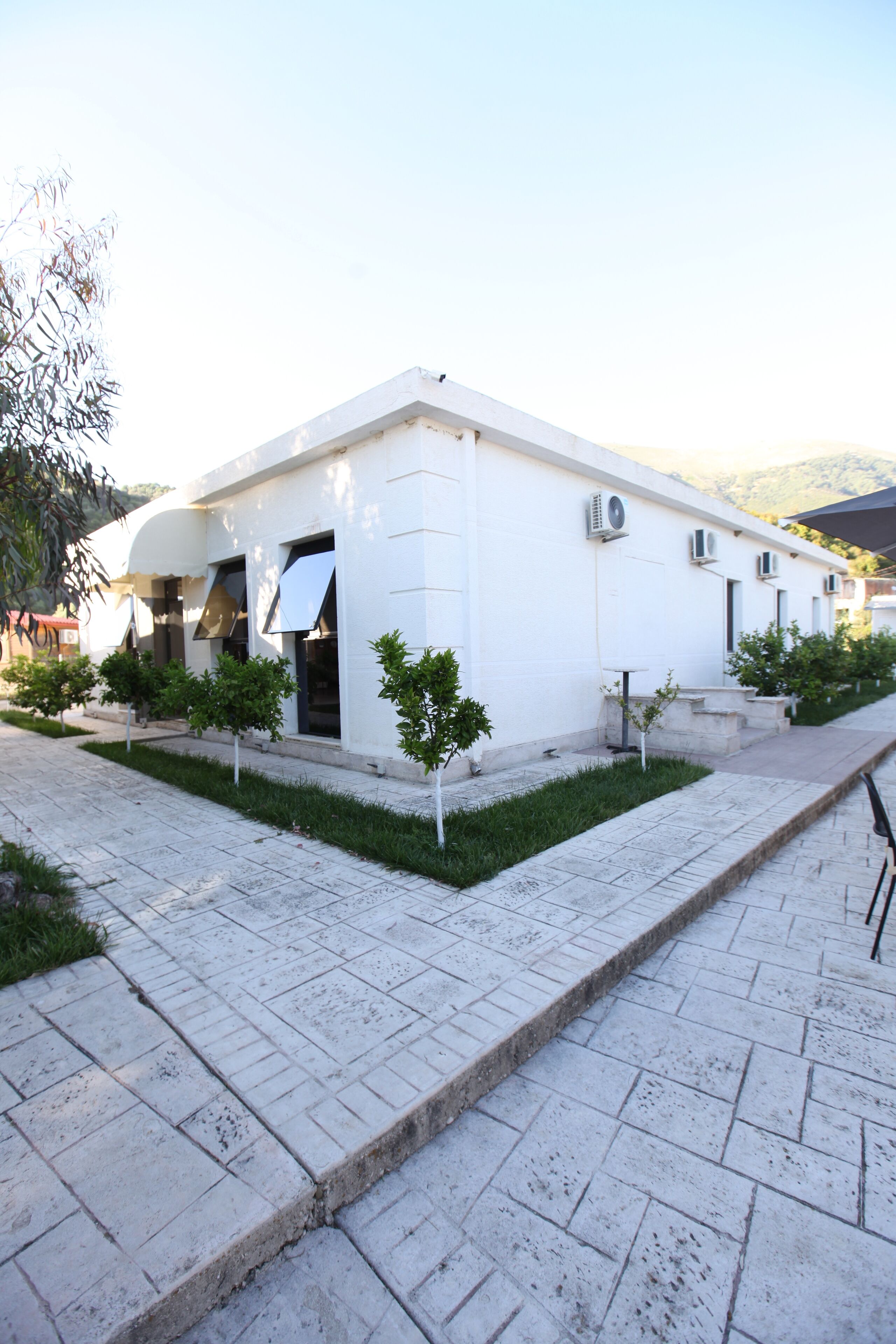 Um edifício branco com telhado plano, aparelhos de ar condicionado e uma pequena área de jardim com árvores jovens.