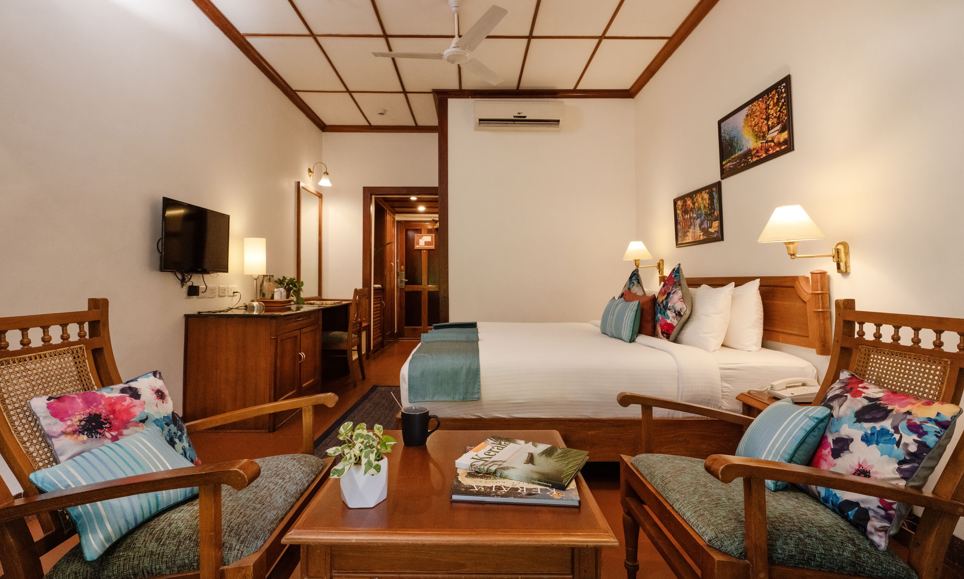 Quarto de hotel com uma cama, duas cadeiras de madeira, uma mesa pequena com uma planta e livros, uma televisão e uma porta que dá para outro quarto.