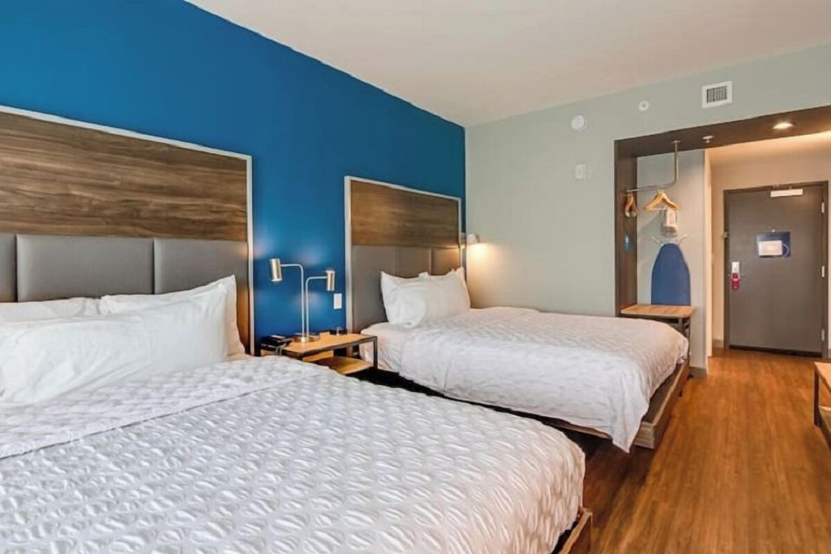 Quarto de hotel com duas camas, uma cabeceira de madeira, uma parede azul de destaque, piso de madeira e uma porta com porta-roupas.