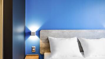 Quarto de hotel com parede azul, mesa de cabeceira de madeira, cama com roupa branca e luminária fixa na parede.