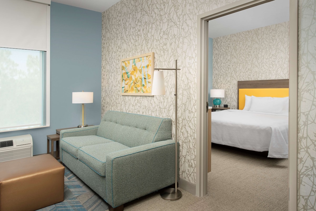 Quarto de hotel com sofá, mesa de cabeceira com abajur, cama com cabeceira e papel de parede estampado.