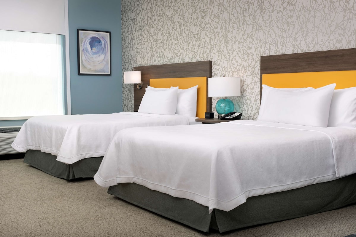 Quarto de hotel com duas camas, cada uma com roupa de cama branca e cabeceira de madeira. Há um globo de parede e um quadro na parede.