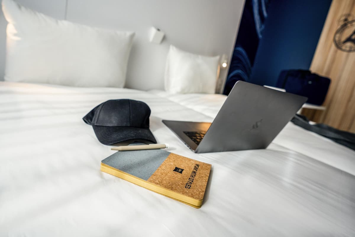Uma cama com um laptop, um boné e um caderno em cima.