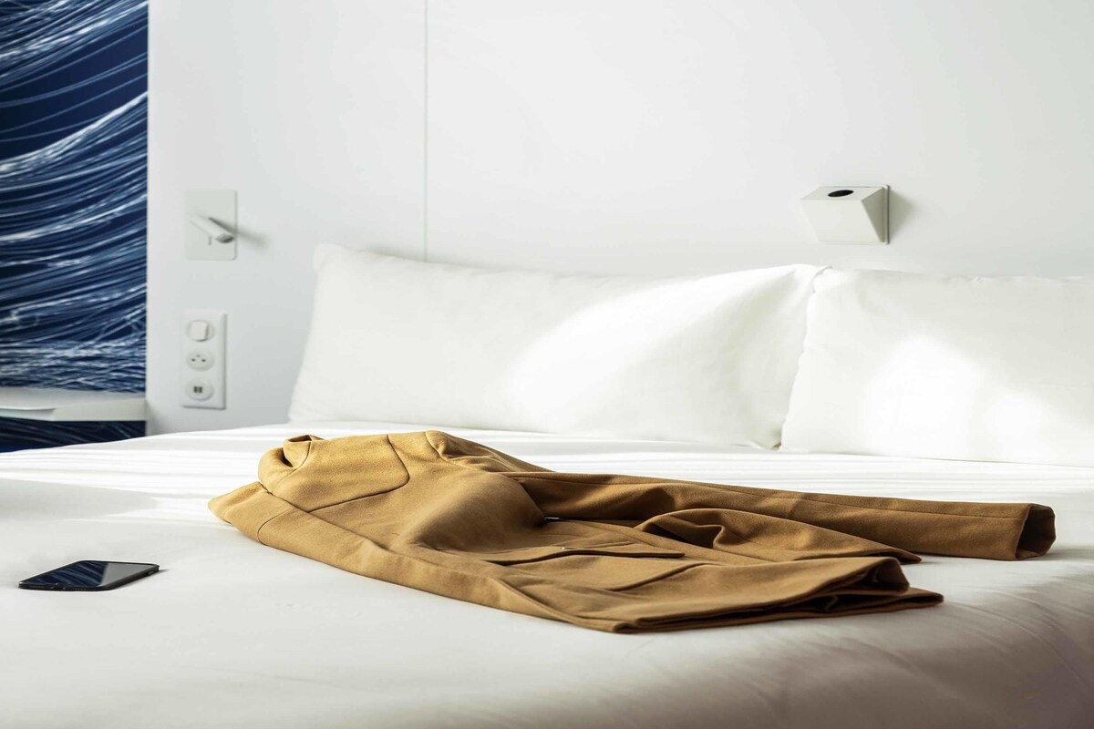 Cama bem arrumada com lençóis brancos, um smartphone sobre a cama e uma peça de roupa castanha na cama.