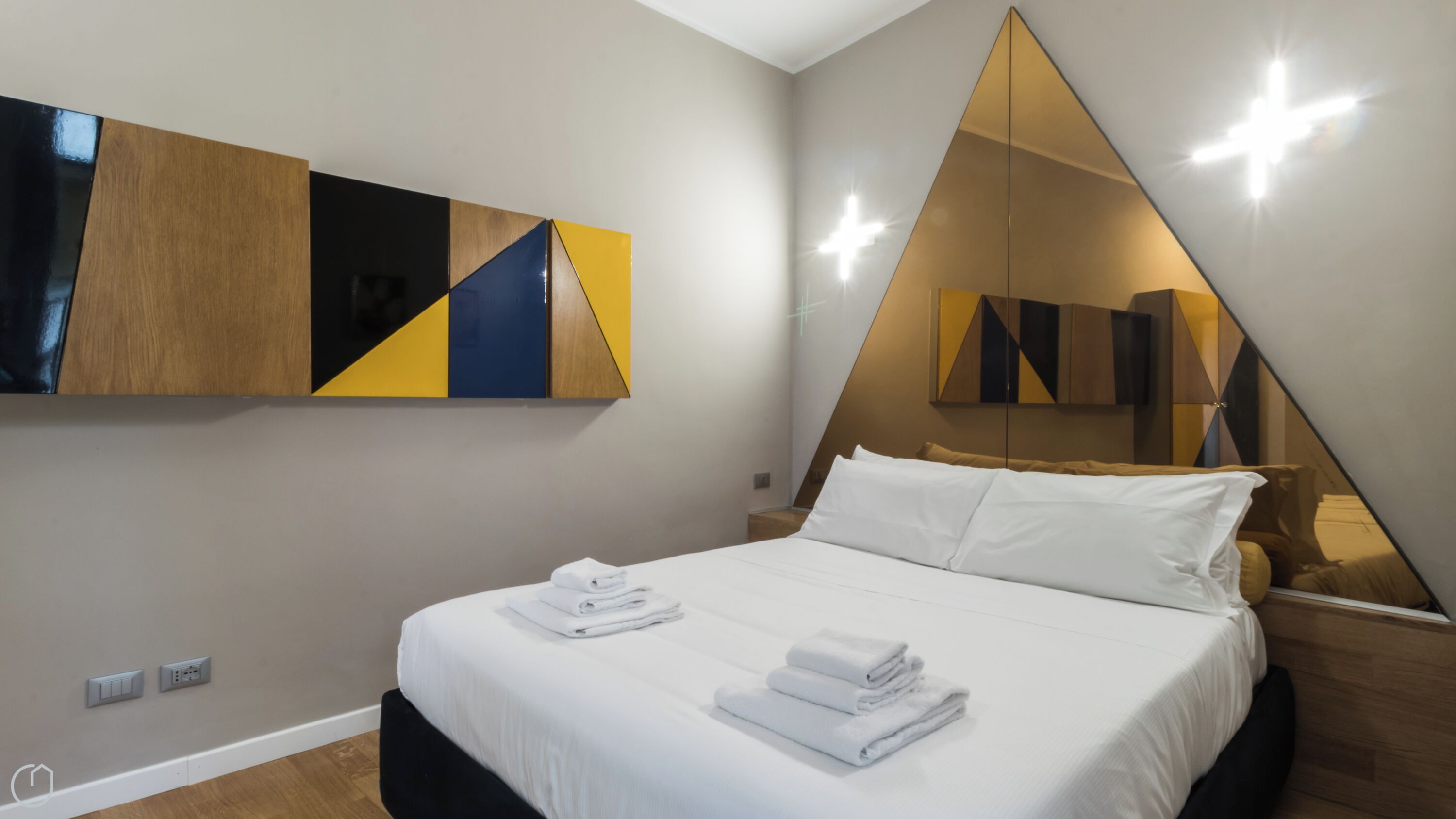 Quarto moderno com cama, roupa de cama branca e uma obra de arte geométrica na parede.