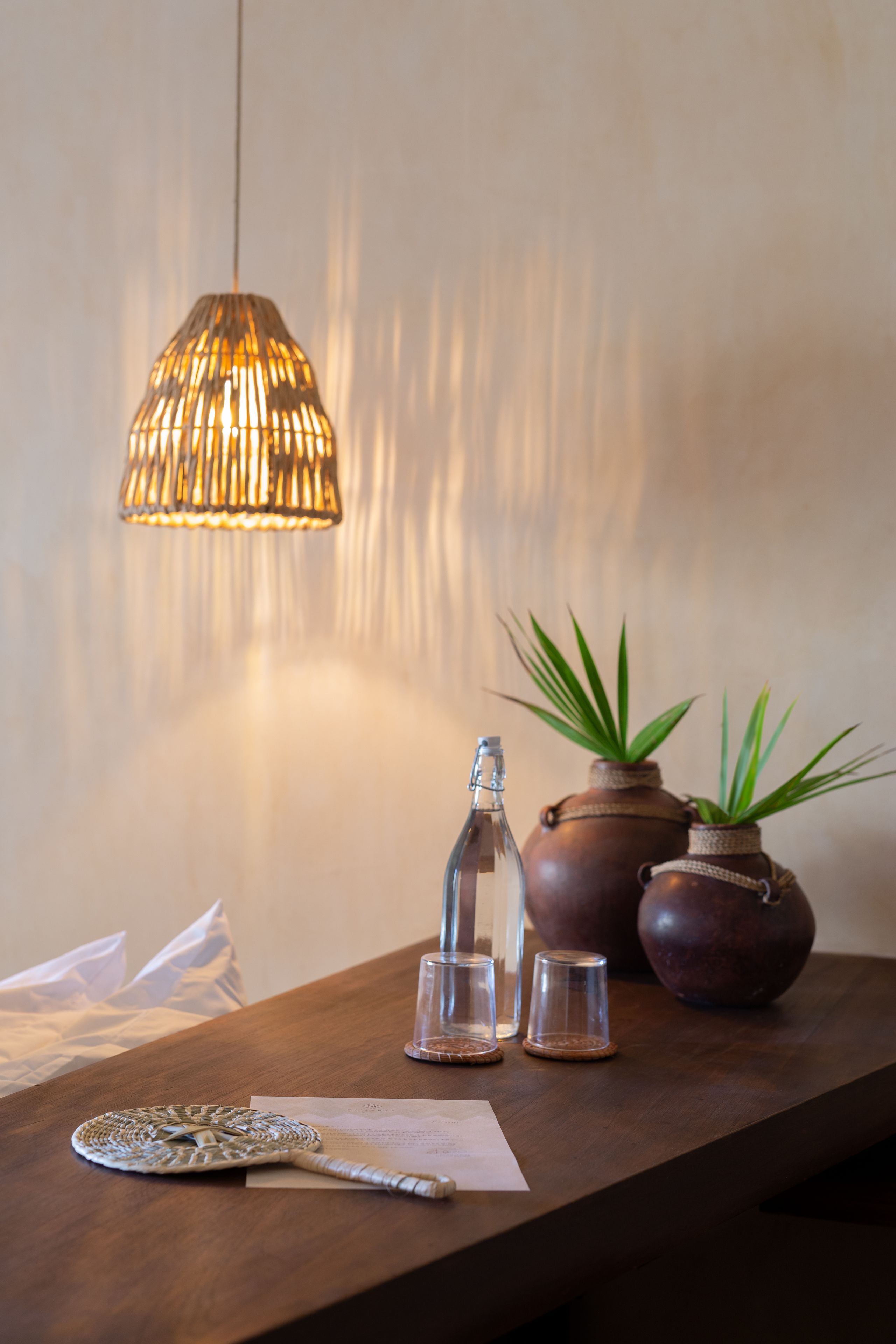 Uma mesa de madeira com uma luminária pendente trançada, uma garrafa transparente, duas taças pequenas, um prato decorativo e duas plantas em vasos.